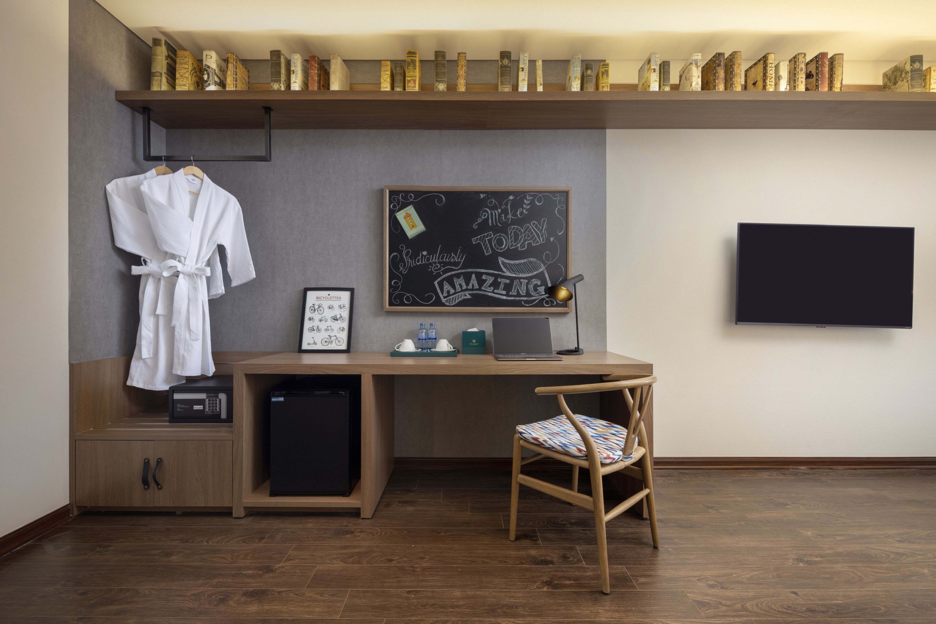 Quarto com escrivaninha de madeira, uma cadeira, um roupão pendurado, estante de livros, quadro negro com escrita, televisão e um pequeno frigorífico.
