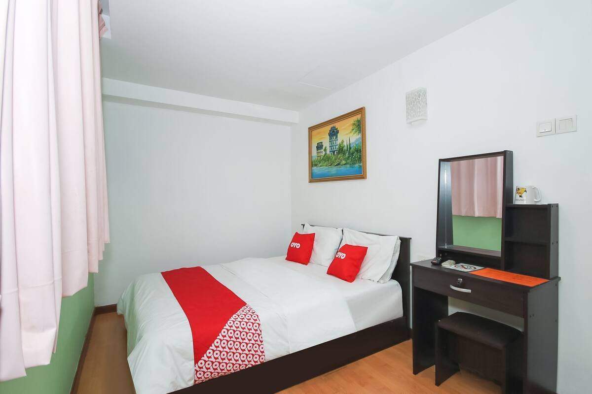 Quarto de hotel com cama, escrivaninha, cadeira, quadro na parede e janela com cortinas.
