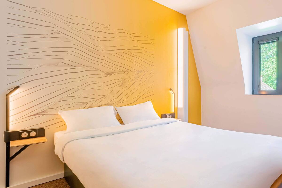 Cama bem arrumada com lençóis brancos, luminária fixada na parede e mesa de cabeceira com rádio.