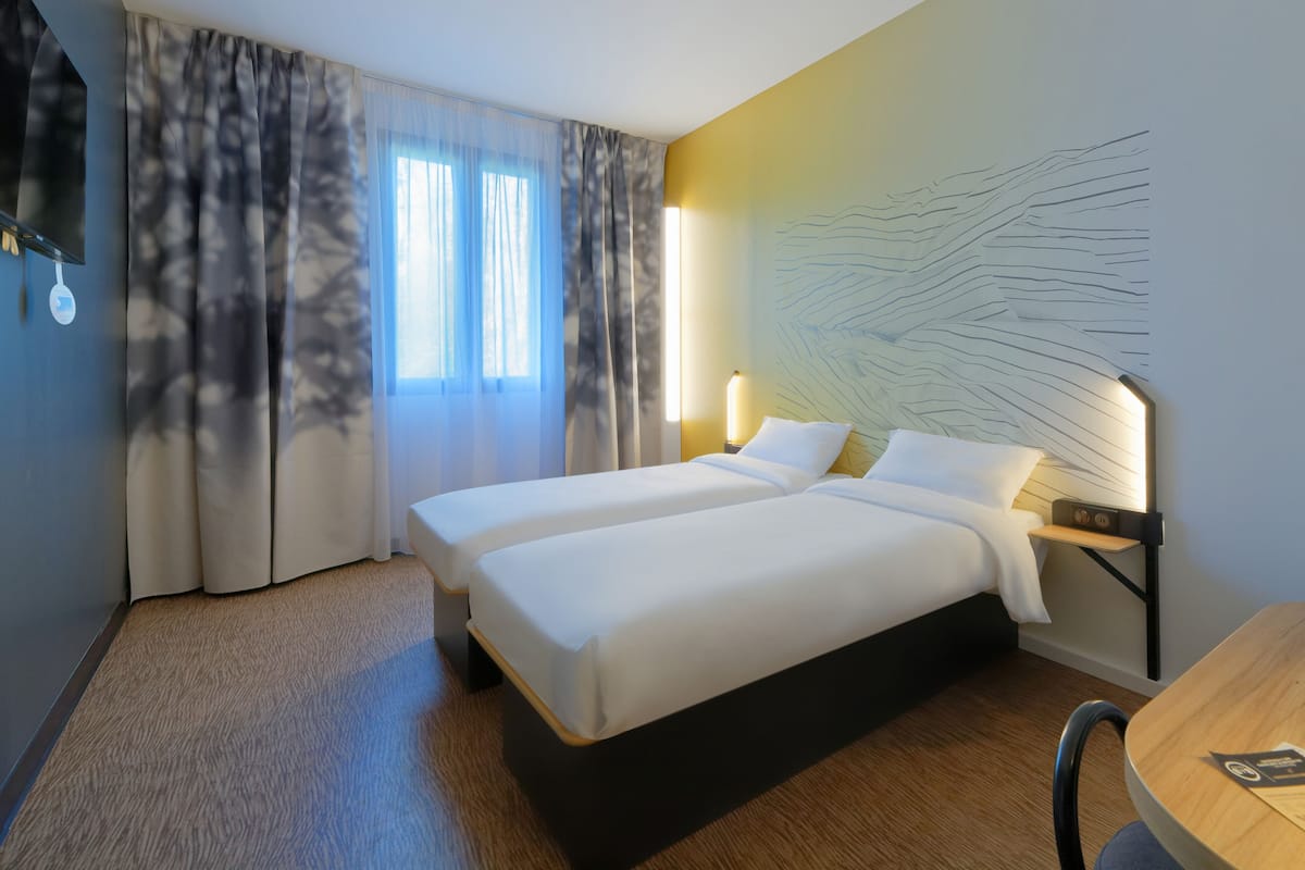 Quarto de hotel com duas camas, uma secretária de madeira, televisão na parede e luminária moderna.