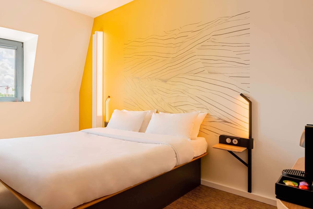 Quarto de hotel com uma cama, uma mesinha de cabeceira, uma luminária na parede e uma janela.