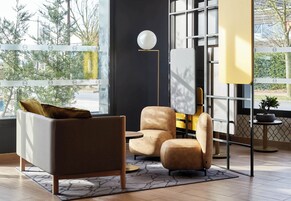 Uma sala de estar moderna com sofá, poltrona e apoio para os pés, uma luminária de chão e vista através de grandes janelas.