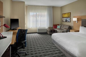 Quarto de hotel com cama, escrivaninha, cadeira, televisão, ar condicionado e janela com cortinas.