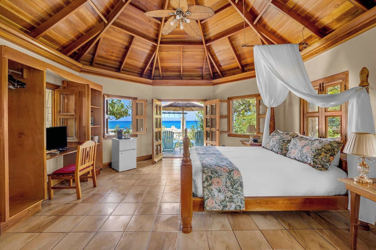 Quarto com teto de madeira, cama com colcha florida, escrivaninha com computador e vista para o mar.