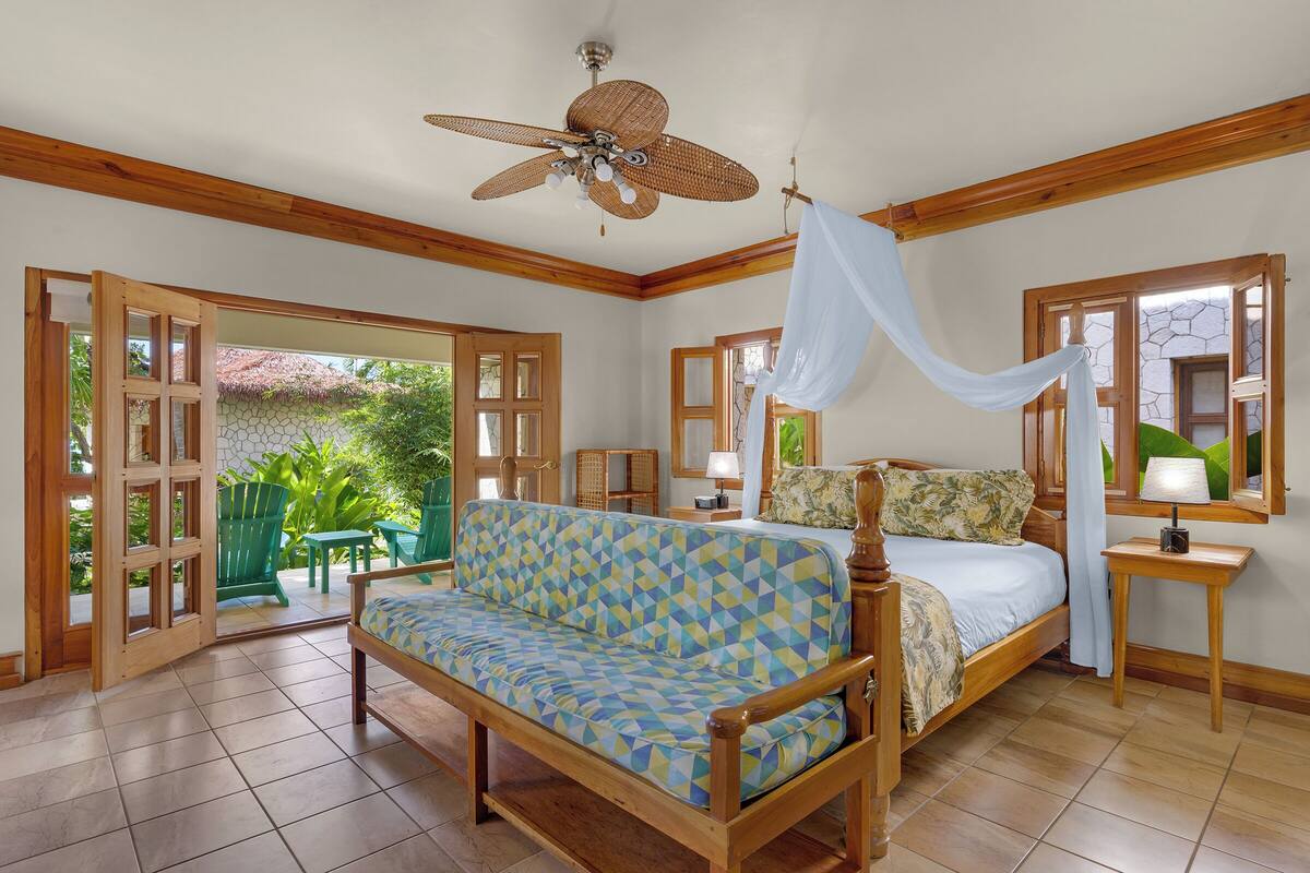 Quarto com cama de madeira, sofá estampado, ventilador de teto e vista para o verde lá fora.