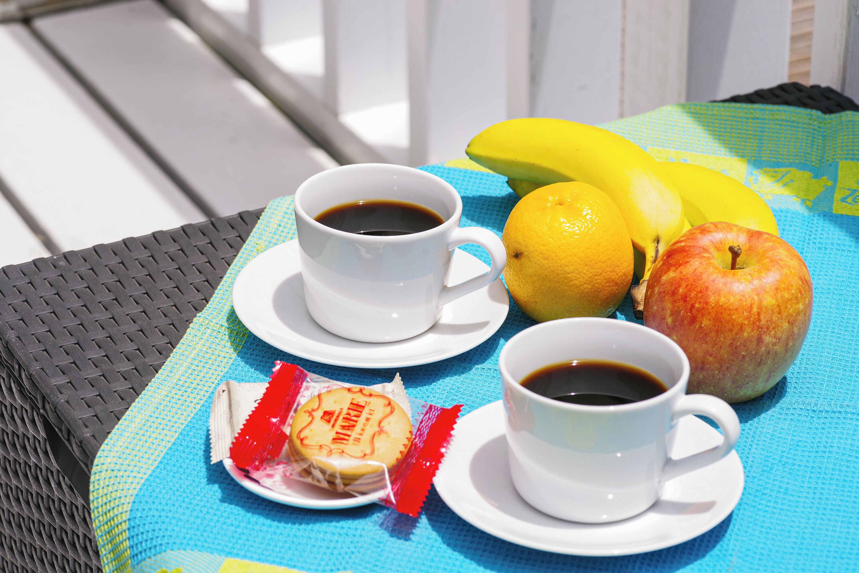Uma mesa com duas chávenas de café, uma banana, uma maçã e um limão sobre um pano de mesa azul.