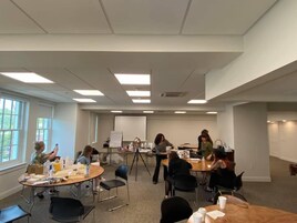 Sala de conferências com mesas redondas, cadeiras, tela de projeção e quadro branco.