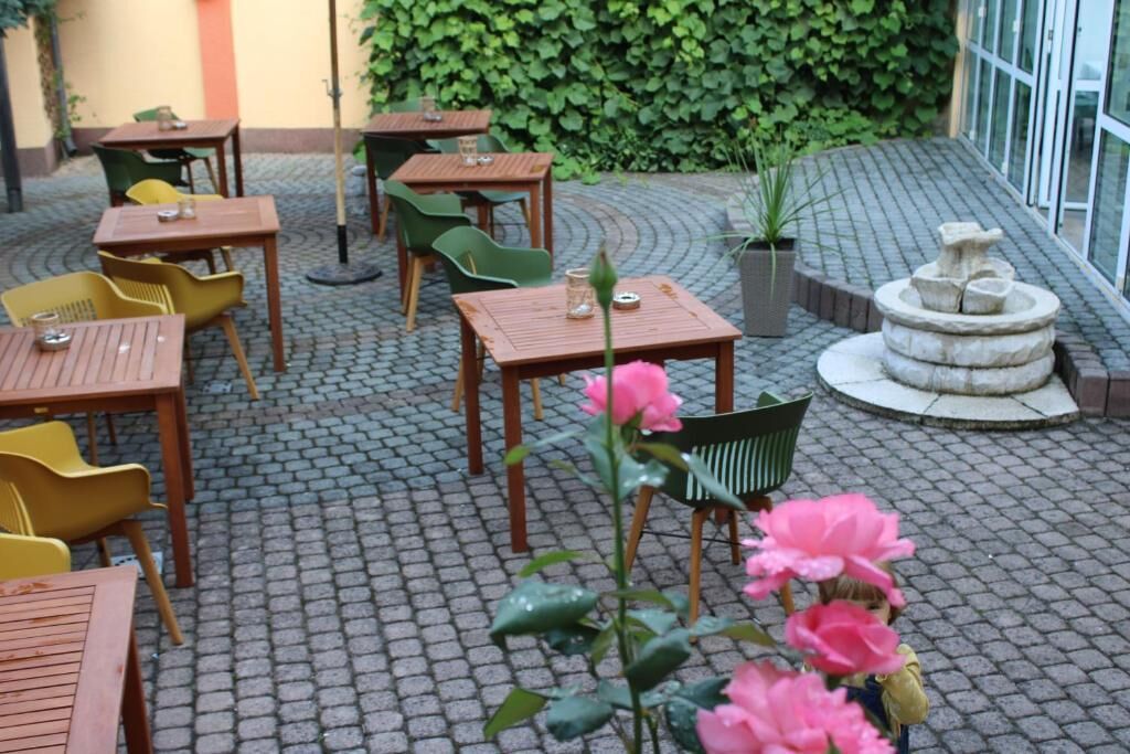Uma área de terraço ao ar livre com mesas e cadeiras de madeira, uma pequena fonte e uma sebe verde ao fundo.