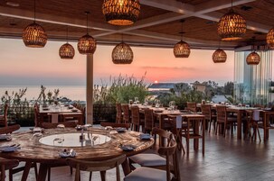 Restaurante com mesas redondas e luminárias de vime suspensas, com vista panorâmica para o oceano e as colinas.