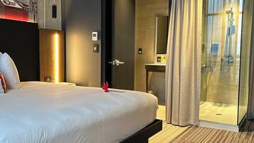 Quarto de hotel moderno com uma cama grande, uma obra de arte urbana, box de vidro para o chuveiro e vista para a cidade pela porta de vidro.