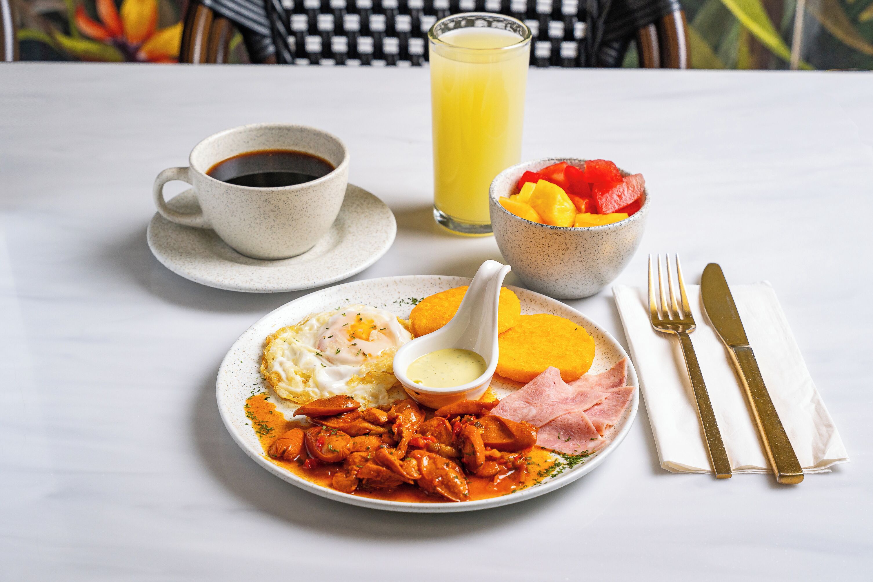 Prato de café da manhã com ovos, presunto e molho, uma xícara de café, um copo de suco de laranja e uma tigela de frutas.