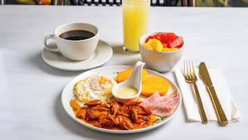 Prato de café da manhã com ovos, presunto e molho, uma xícara de café, um copo de suco de laranja e uma tigela de frutas.
