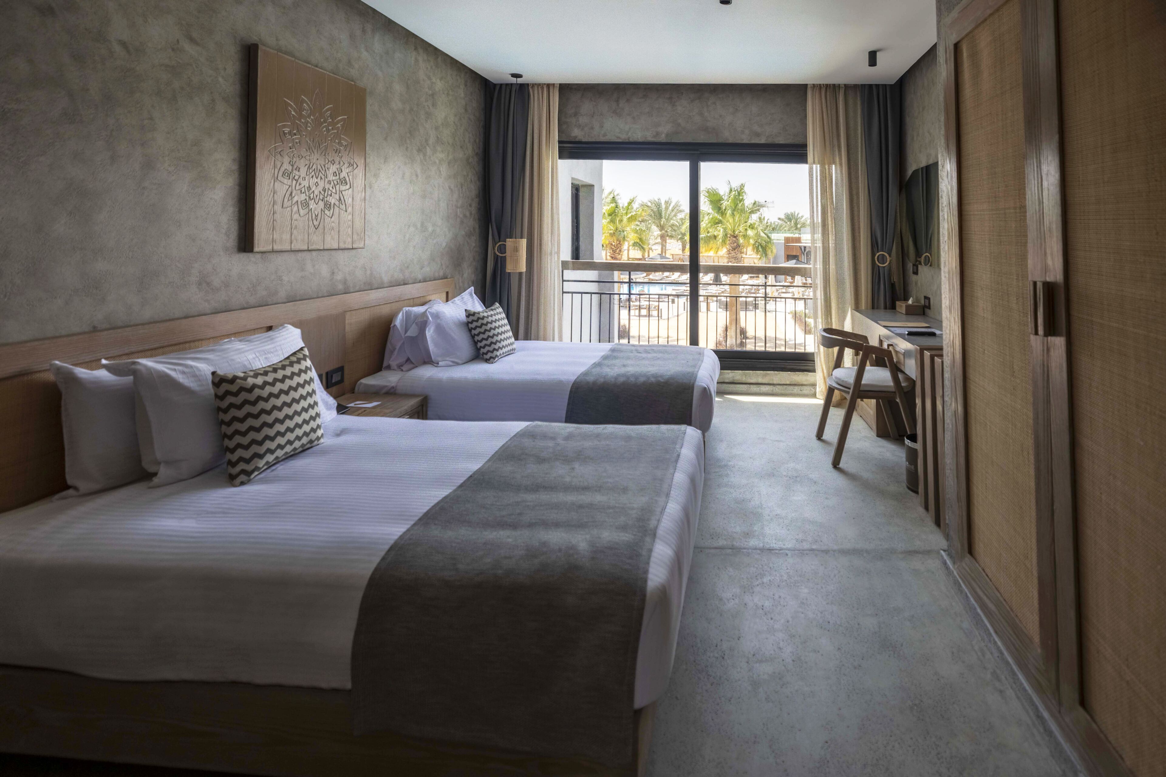 Quarto de hotel com duas camas, uma escrivaninha, uma cadeira, televisão e varanda com vista para palmeiras.