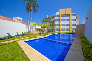 Uma piscina com borda em azulejo, cercada por grama e palmeiras, com edifícios modernos ao fundo.