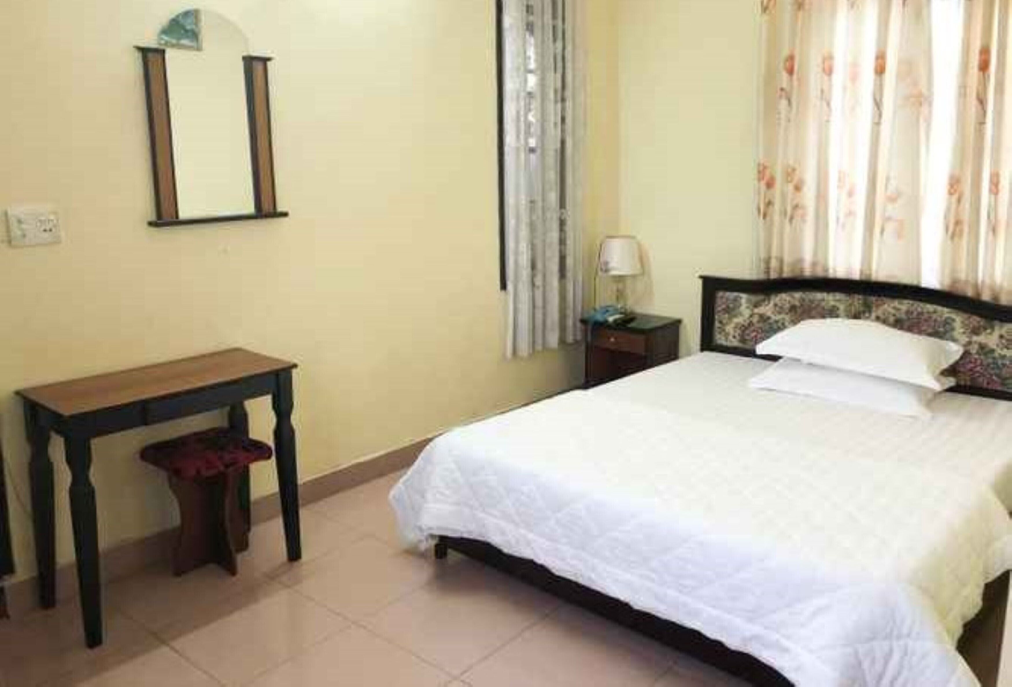 Quarto com cama, mesa pequena, cadeira, mesa de cabeceira, abajur e espelho.