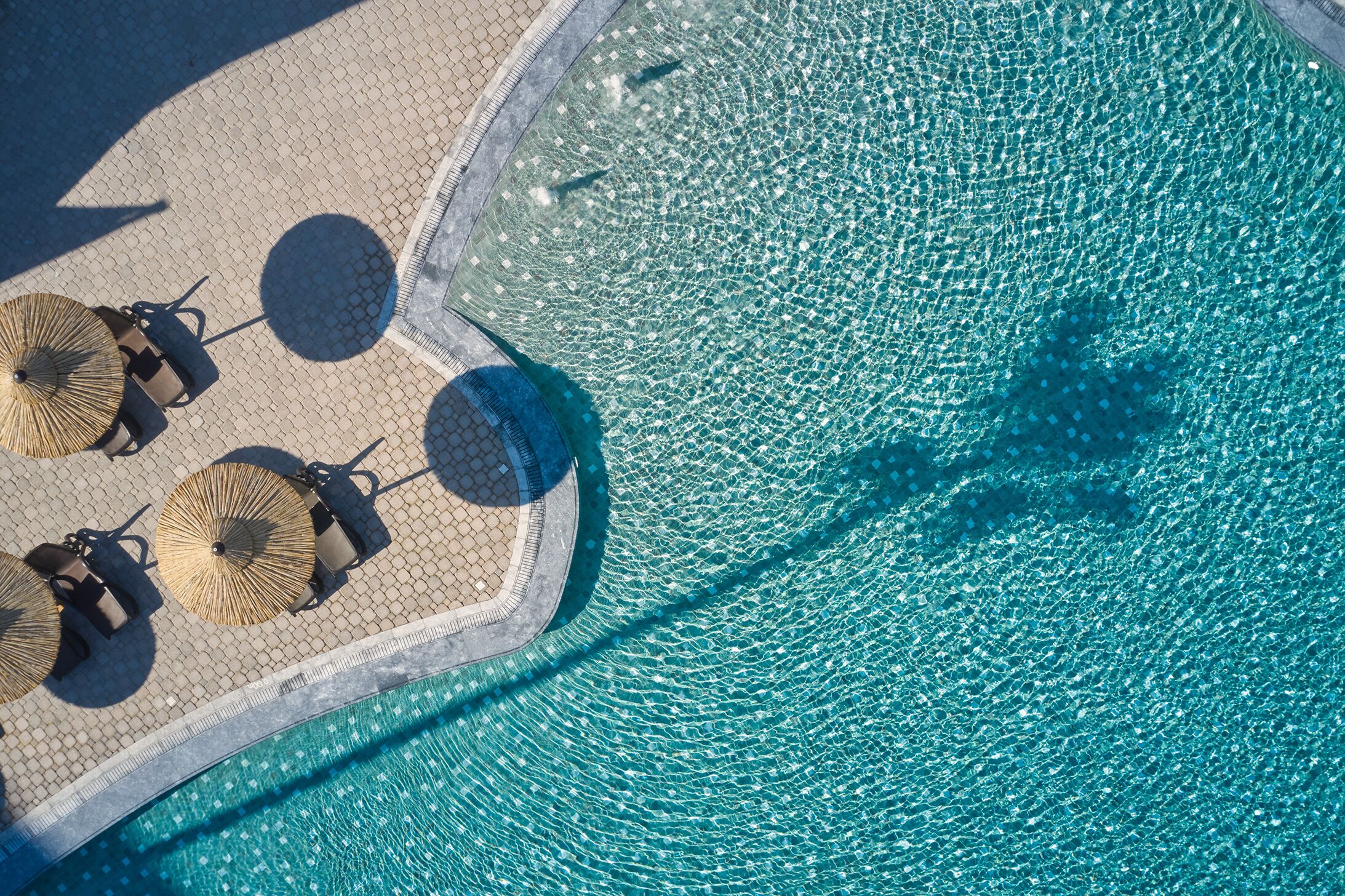 Vista aérea de uma piscina com água cristalina, rodeada por um deck revestido com guarda-sóis e espreguiçadeiras.