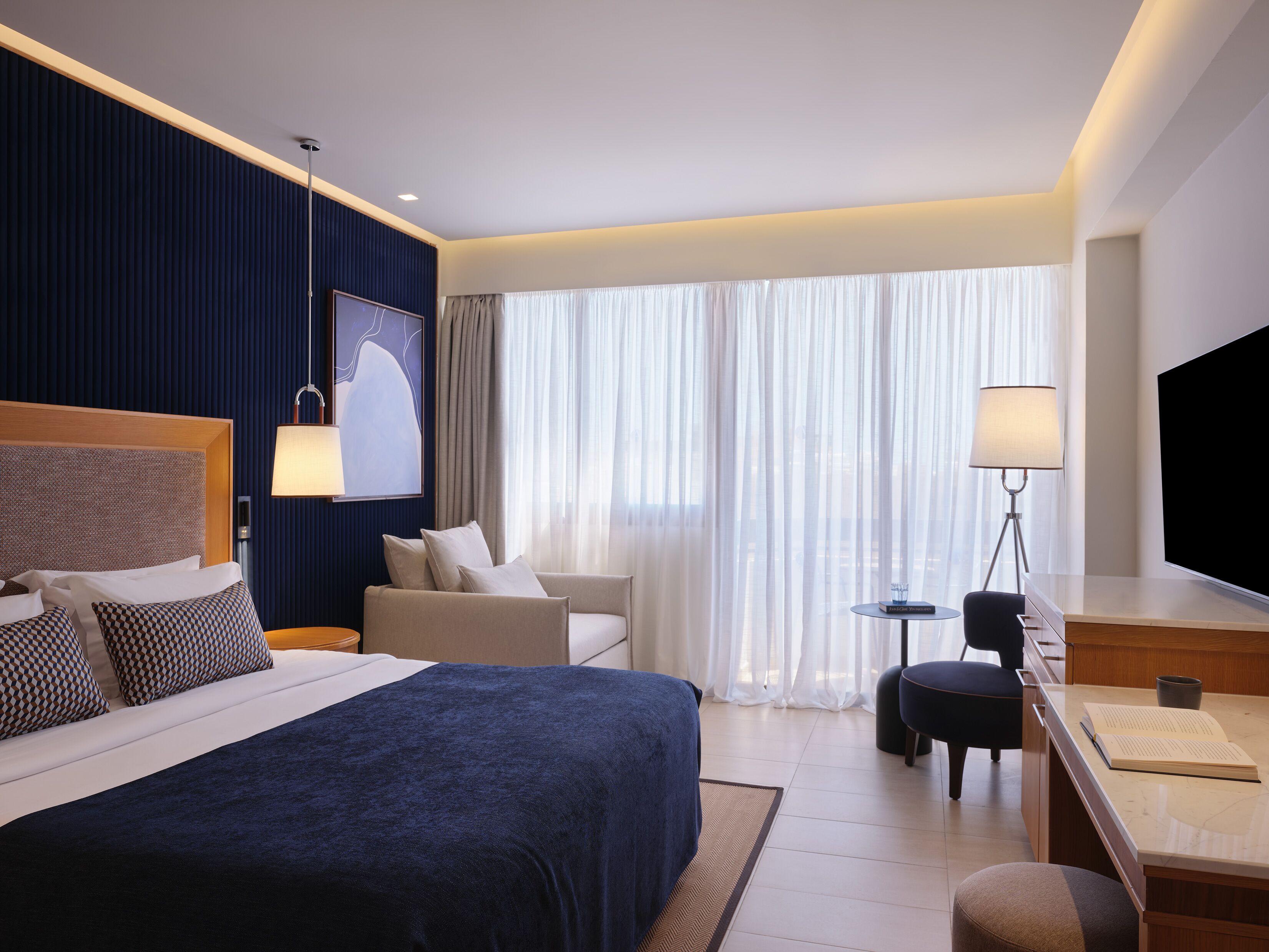 Quarto de hotel com uma cama grande, uma escrivaninha com luminária, televisão, uma cadeira e janela com cortinas.