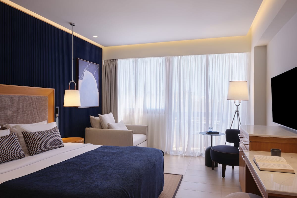 Quarto de hotel com uma cama grande, uma escrivaninha com luminária, televisão, uma cadeira e janela com cortinas.