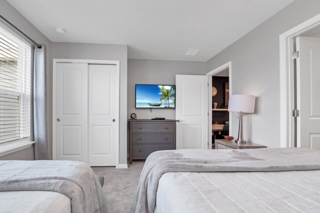 Quarto com duas camas, uma televisão sobre um penteadeira, uma luminária e uma porta que dá para outro cômodo.