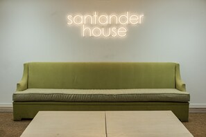 Um sofá verde com uma almofada de cor clara em frente a uma parede com uma placa de neon que diz "santander house".