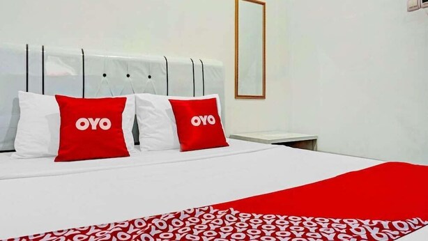Quarto de hotel com uma cama, duas almofadas vermelhas com a palavra 'OYO' e uma cabeceira branca, mesa de cabeceira e um espelho na parede.