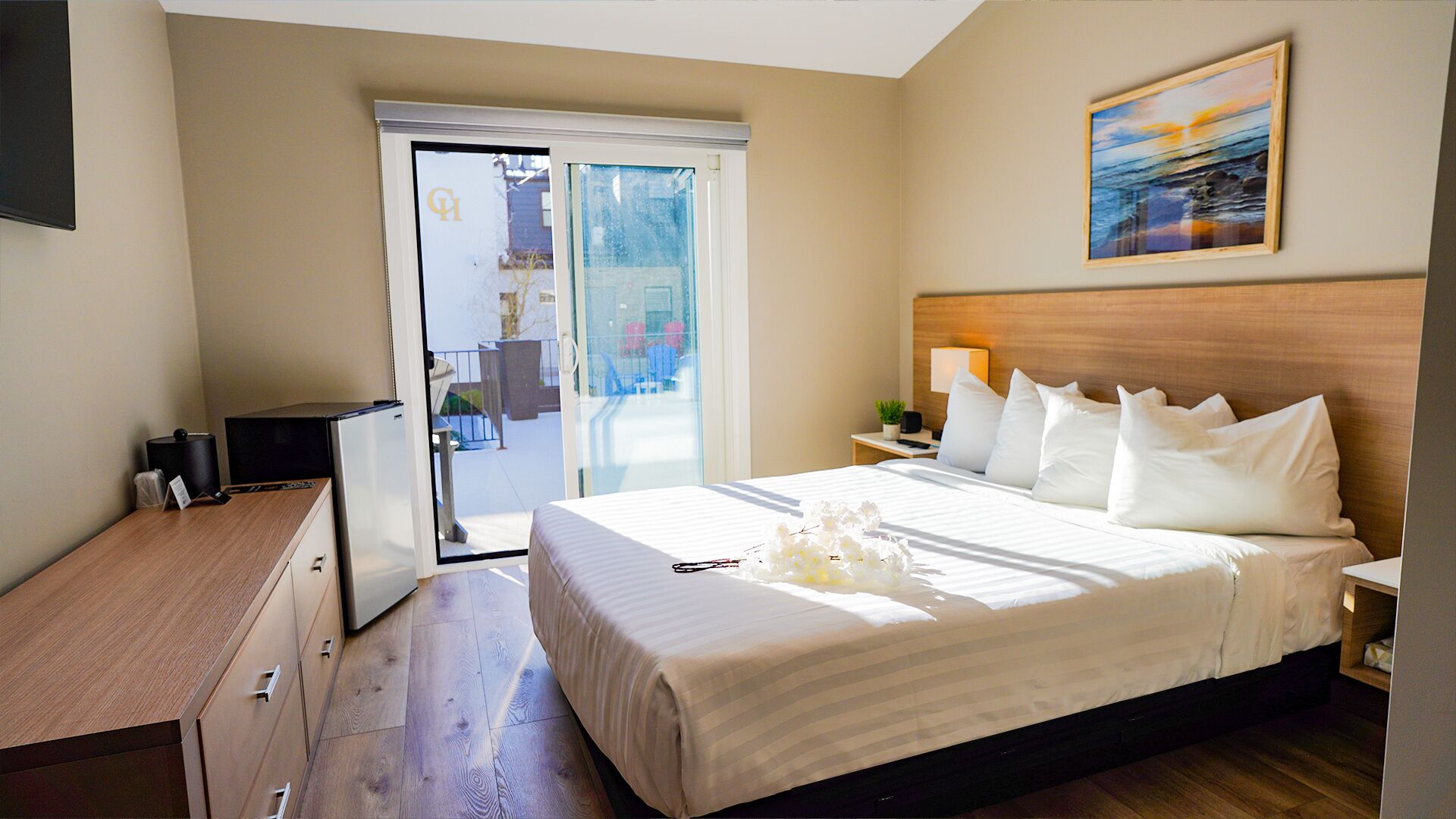 Quarto de hotel com uma cama, uma escrivaninha, um frigorífico, uma mesinha de cabeceira com uma lâmpada e um quadro na parede.