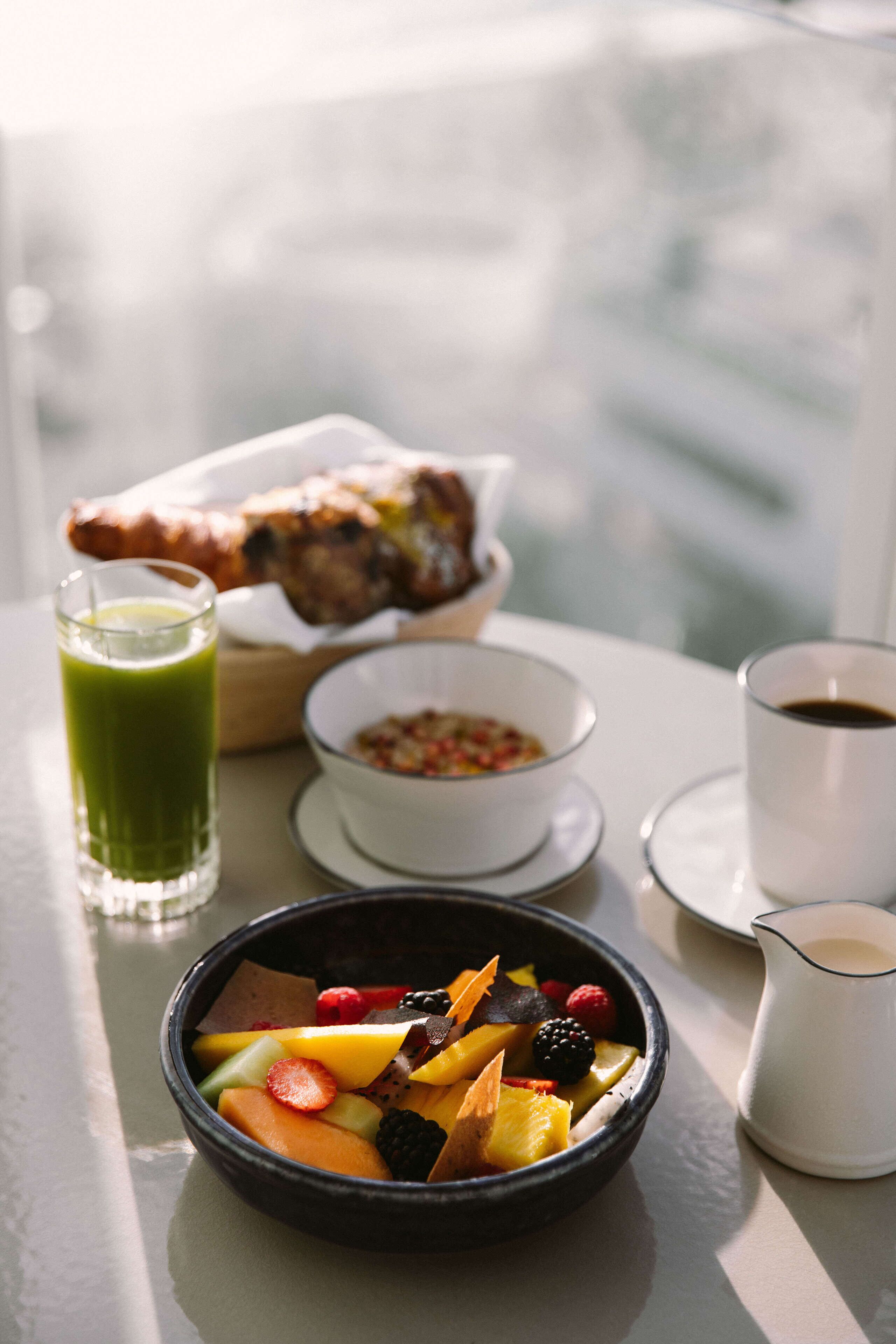 Uma mesa de café da manhã com uma tigela de frutas, um copo de suco verde, uma xícara de café e uma cesta de pão.