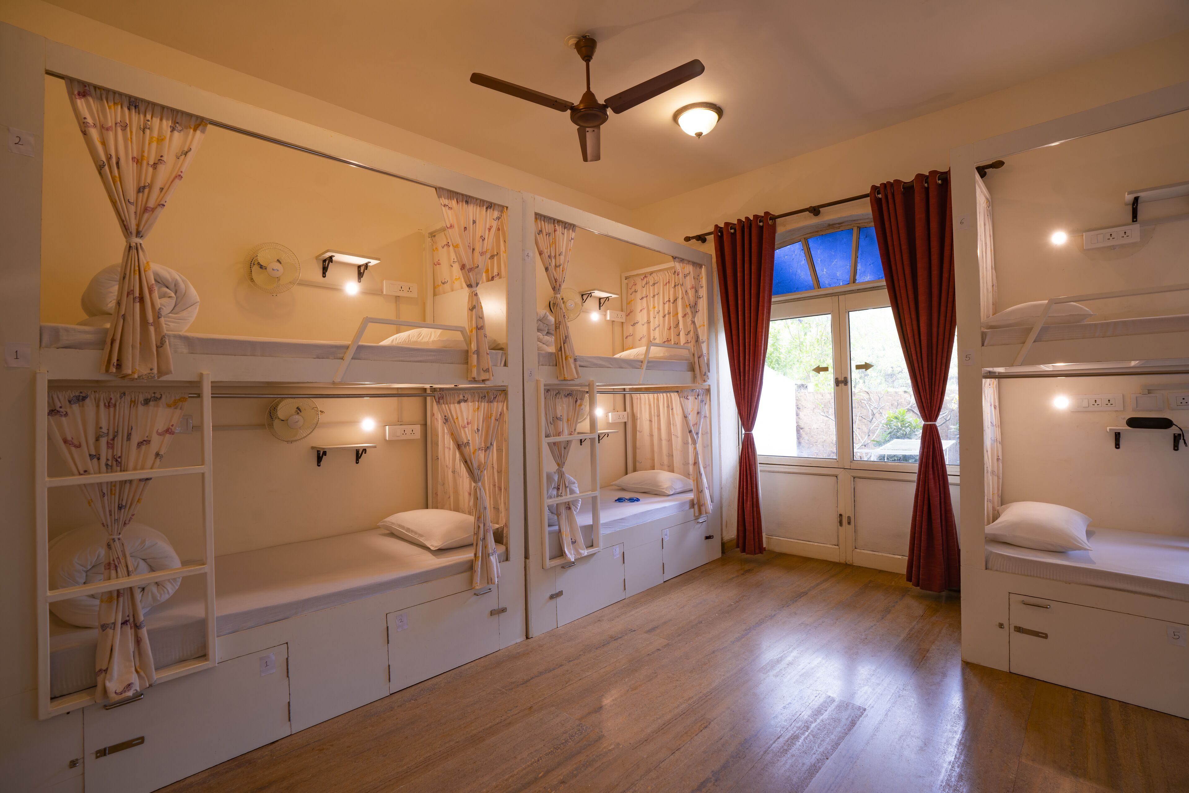 Quarto em dormitório com beliches, ventilador de teto e janela com cortinas.
