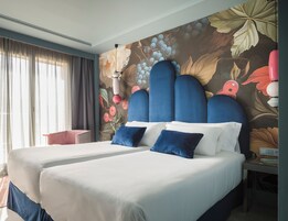 Quarto de hotel com duas camas, um grande mural floral na parede e uma janela com cortinas leves.