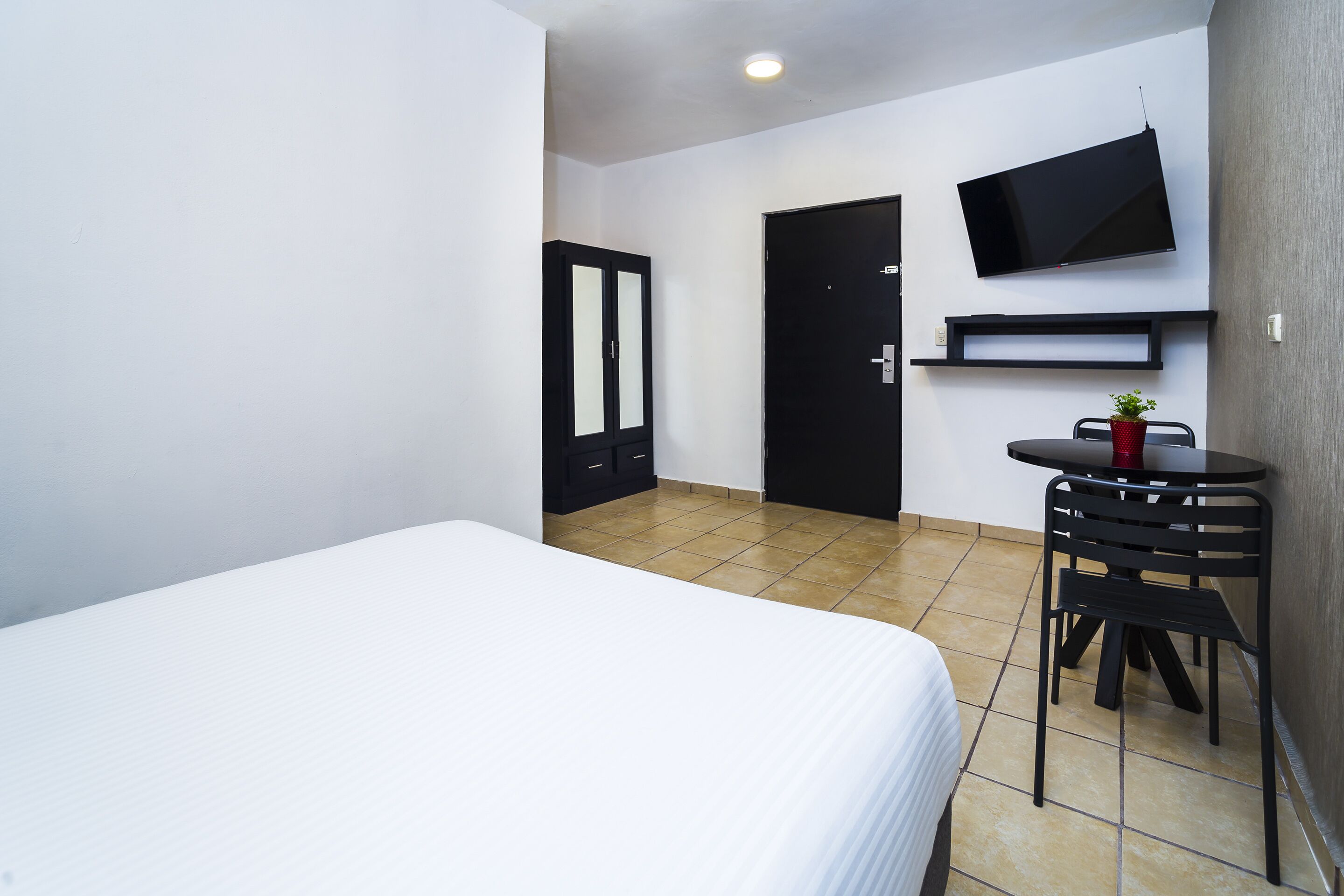Quarto de hotel com uma cama, uma escrivaninha com cadeira, uma televisão na parede e uma porta.
