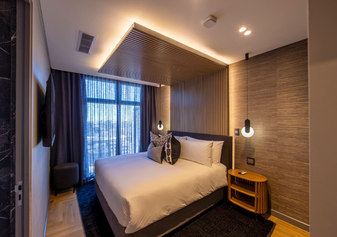 Quarto de hotel moderno com uma cama grande, uma mesa de cabeceira, uma televisão fixada na parede e uma janela com cortinas.