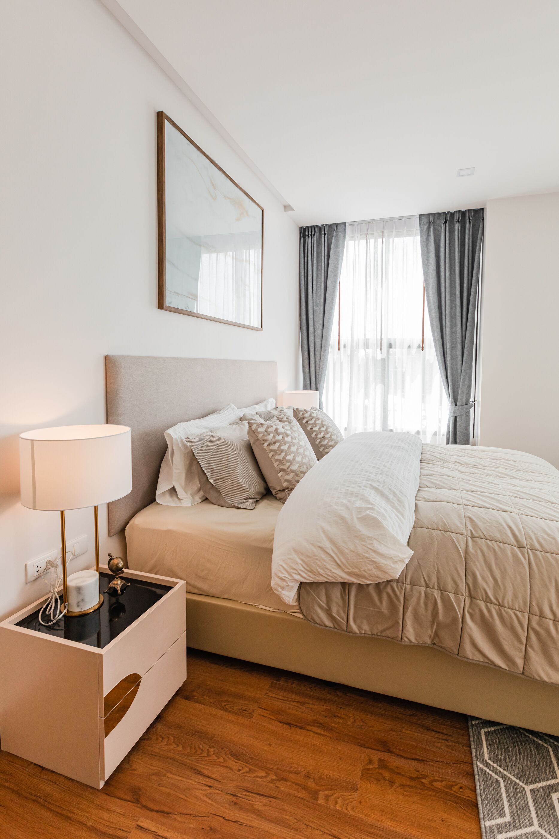 Quarto bem arrumado com cama, mesa de cabeceira, abajur e janela com cortinas.