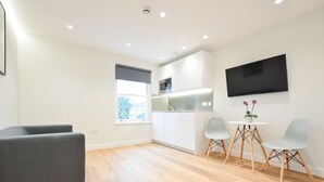 Uma sala de estar moderna com um sofá cinzento, uma mesa de jantar com duas cadeiras, uma televisão de tela plana na parede e uma área de cozinha com armários brancos e um micro-ondas.