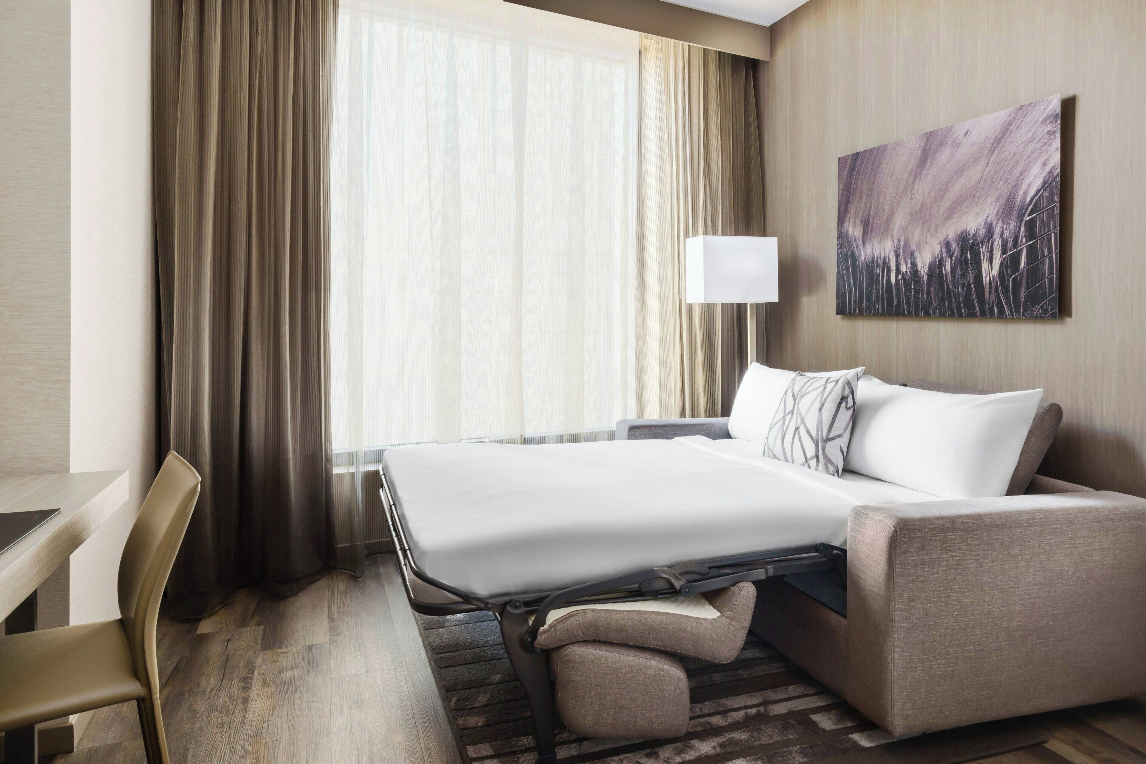 Quarto de hotel com cama, escrivaninha, poltrona, sofá, luminária e um quadro na parede.