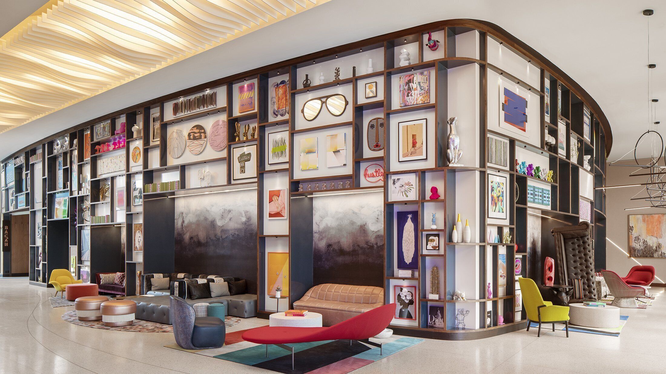 Um interior moderno com uma parede curva exibindo diversas obras de arte e objetos decorativos. Há várias áreas de estar com cadeiras e pufes coloridos.