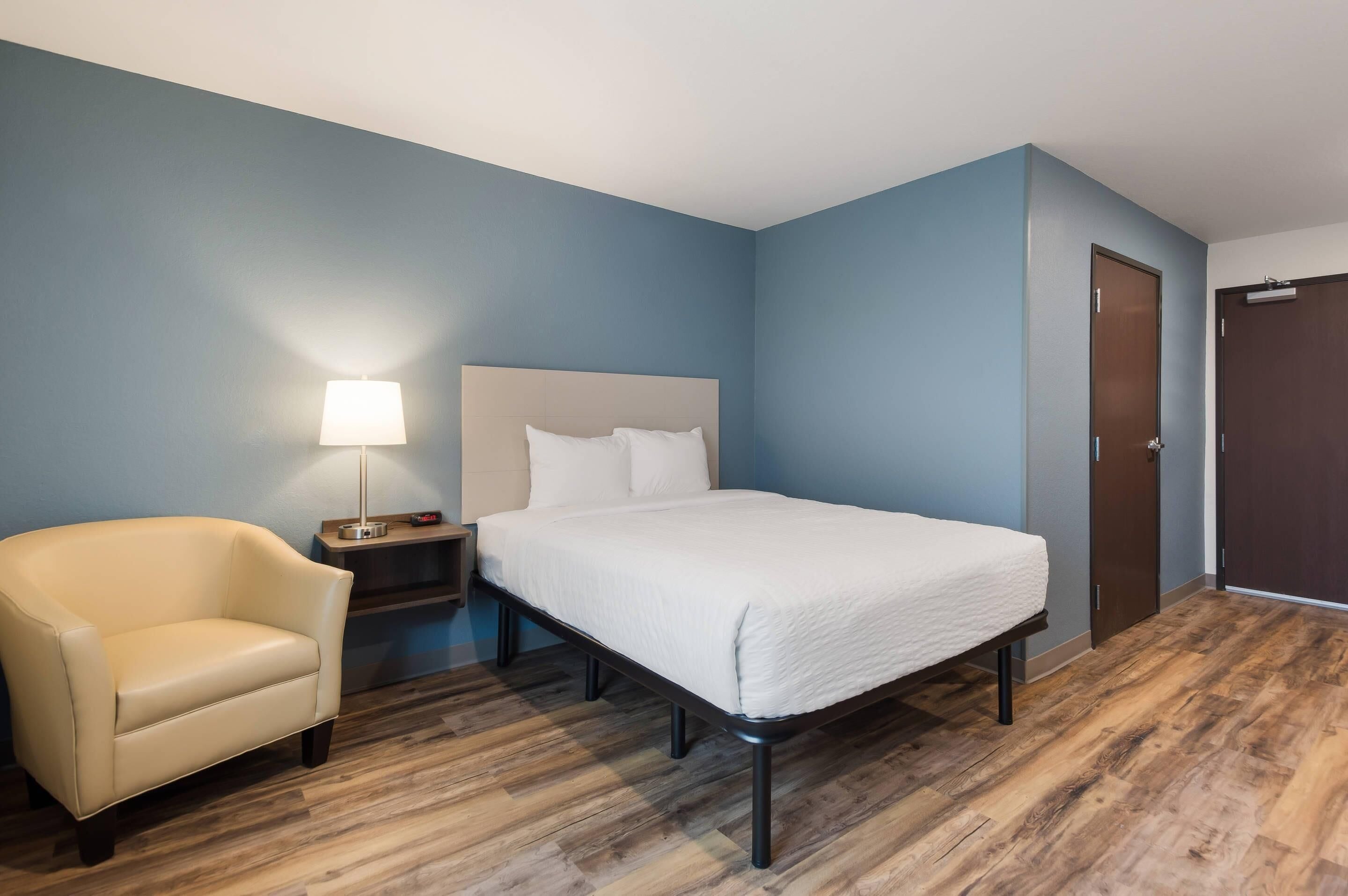Quarto de hotel com cama, cadeira, mesa de cabeceira e abajur.