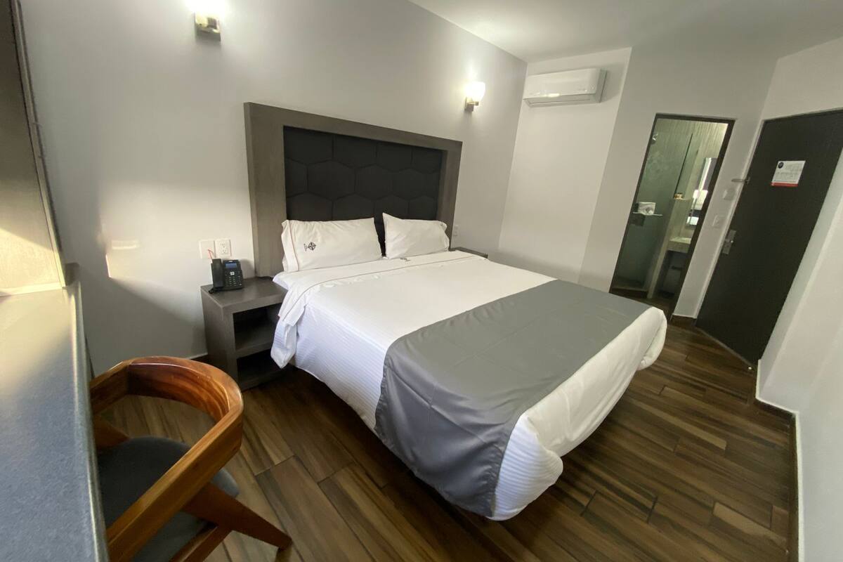 Quarto de hotel com uma cama grande, uma cadeira de madeira, uma mesa de cabeceira com telefone e um ar condicionado na parede.