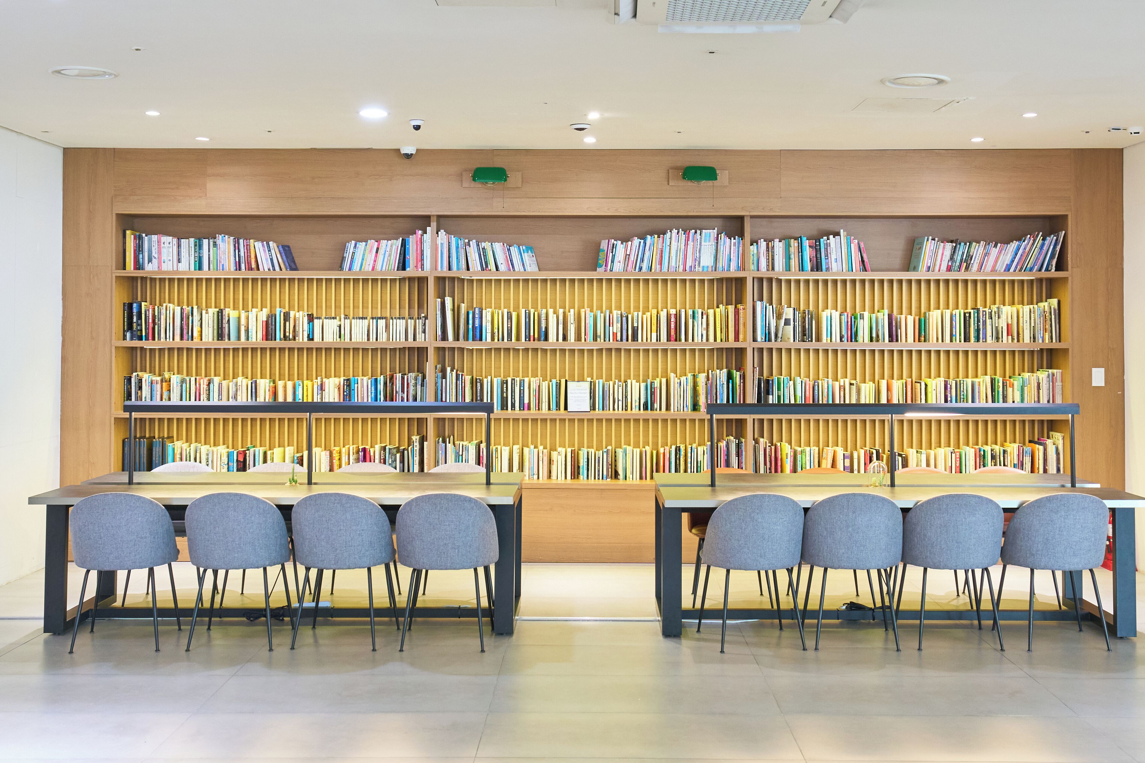 Uma biblioteca com várias estantes, duas mesas com cadeiras e um teto com iluminação embutida.