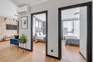 Quarto de hotel moderno com parede de tijolos, televisão de tela plana, mesa de cabeceira, planta e cama com colcha cinza.