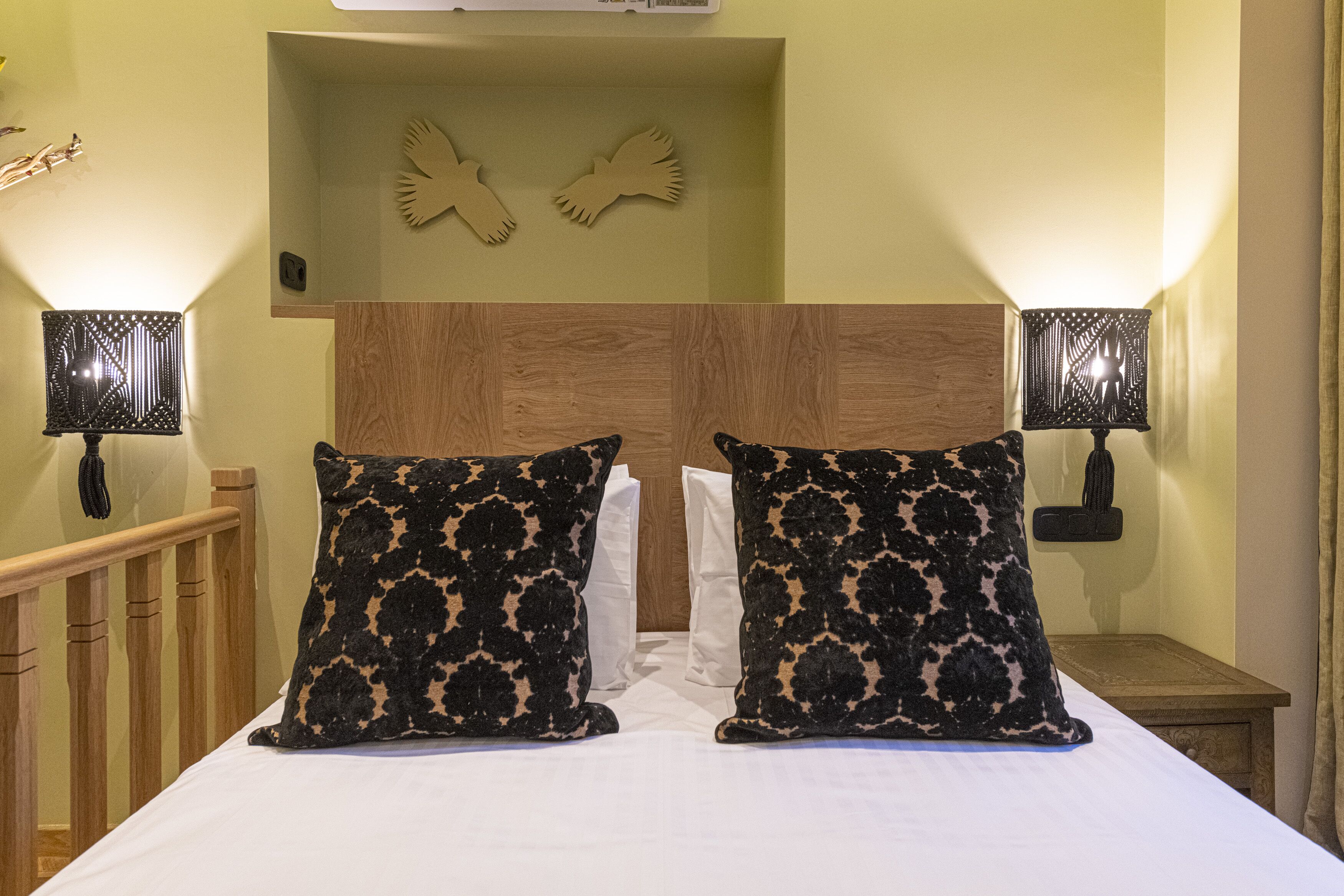 Quarto de hotel com cabeceira de madeira, duas almofadas estampadas em preto e bege, colcha branca e duas luminárias fixadas na parede.