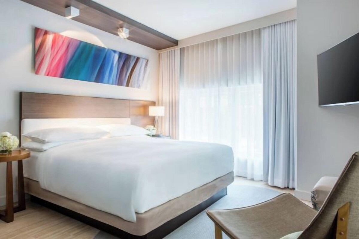 Quarto de hotel com uma cama grande, uma mesa de cabeceira com abajur, uma TV fixada na parede e uma janela com cortinas.