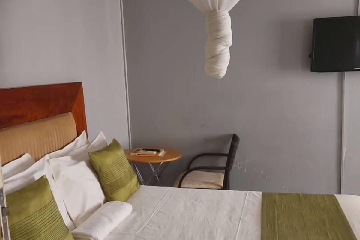 Quarto de hotel com uma cama, uma cadeira, uma mesinha e uma televisão na parede.