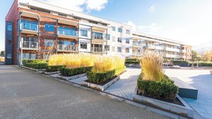 Um edifício residencial moderno de vários andares, com varandas, entrada pavimentada e áreas ajardinadas com gramíneas altas.