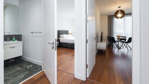 Quarto de hotel moderno com piso de madeira, banheiro branco com toucador de mármore, cama com roupa de cama branca e área de jantar com mesa e cadeiras.