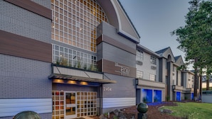Hotel com design moderno, apresentando uma fachada de vidro ampla, entrada bem iluminada e área externa ajardinada com bancos e um espelho d'água.