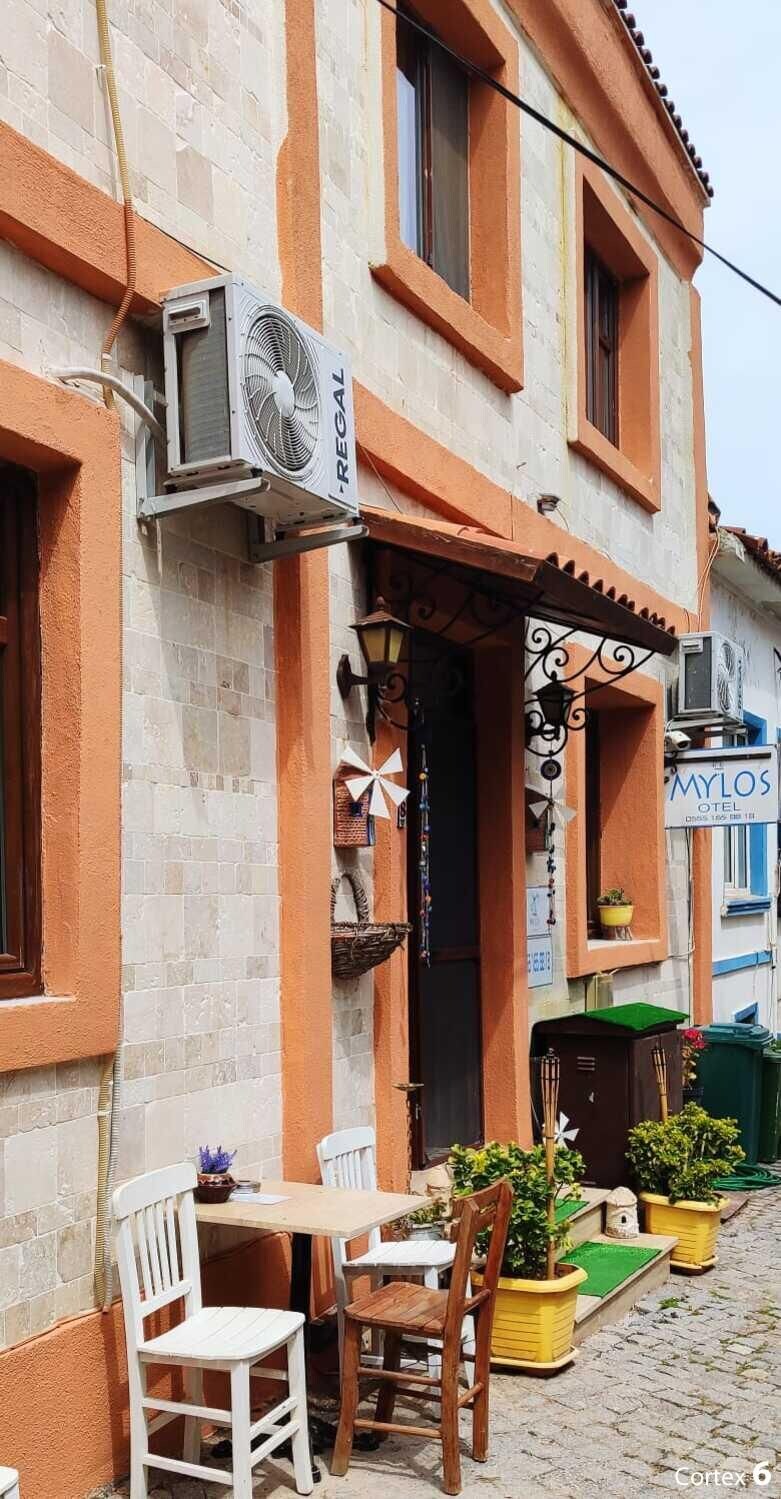Um edifício com fachada em azulejos, um letreiro com a inscrição 'MYLOS HOTEL' e área externa com mesas e cadeiras.