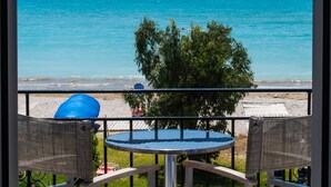 Varanda com cadeiras de vime e uma mesa, com vista para uma praia com água azul cristalina e um veleiro.
