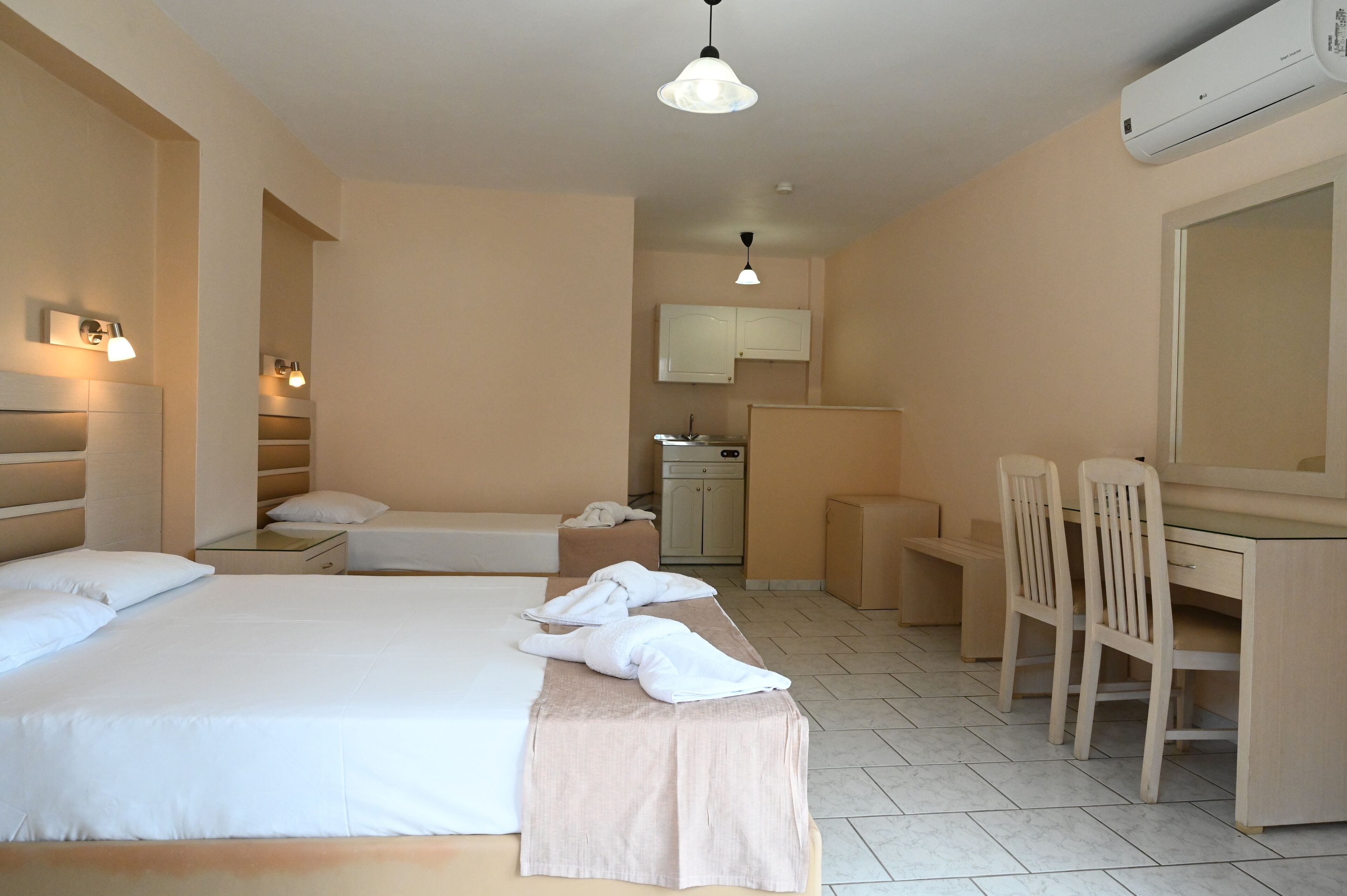 Quarto de hotel com duas camas, uma escrivaninha, uma cadeira, uma kitchenette pequena e um espelho.