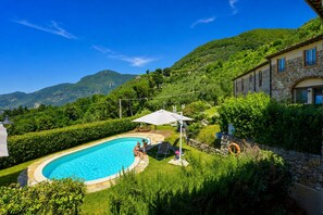 Piscina cercada por vegetação exuberante, um edifício de pedra e um cenário montanhoso.