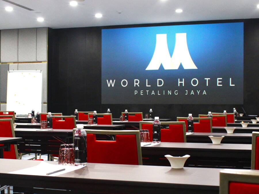 Uma sala de conferências com mesas e cadeiras, uma tela grande exibindo o logotipo do World Hotel Petaling Jaya e uma lousa ao fundo.